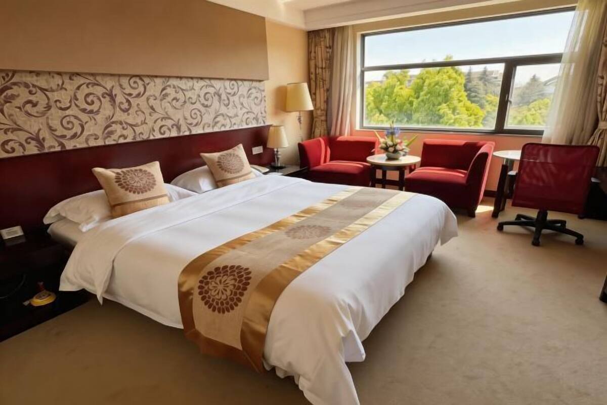 Quarto de hotel com uma cama grande, uma escrivaninha, duas cadeiras e uma janela com vista para árvores.