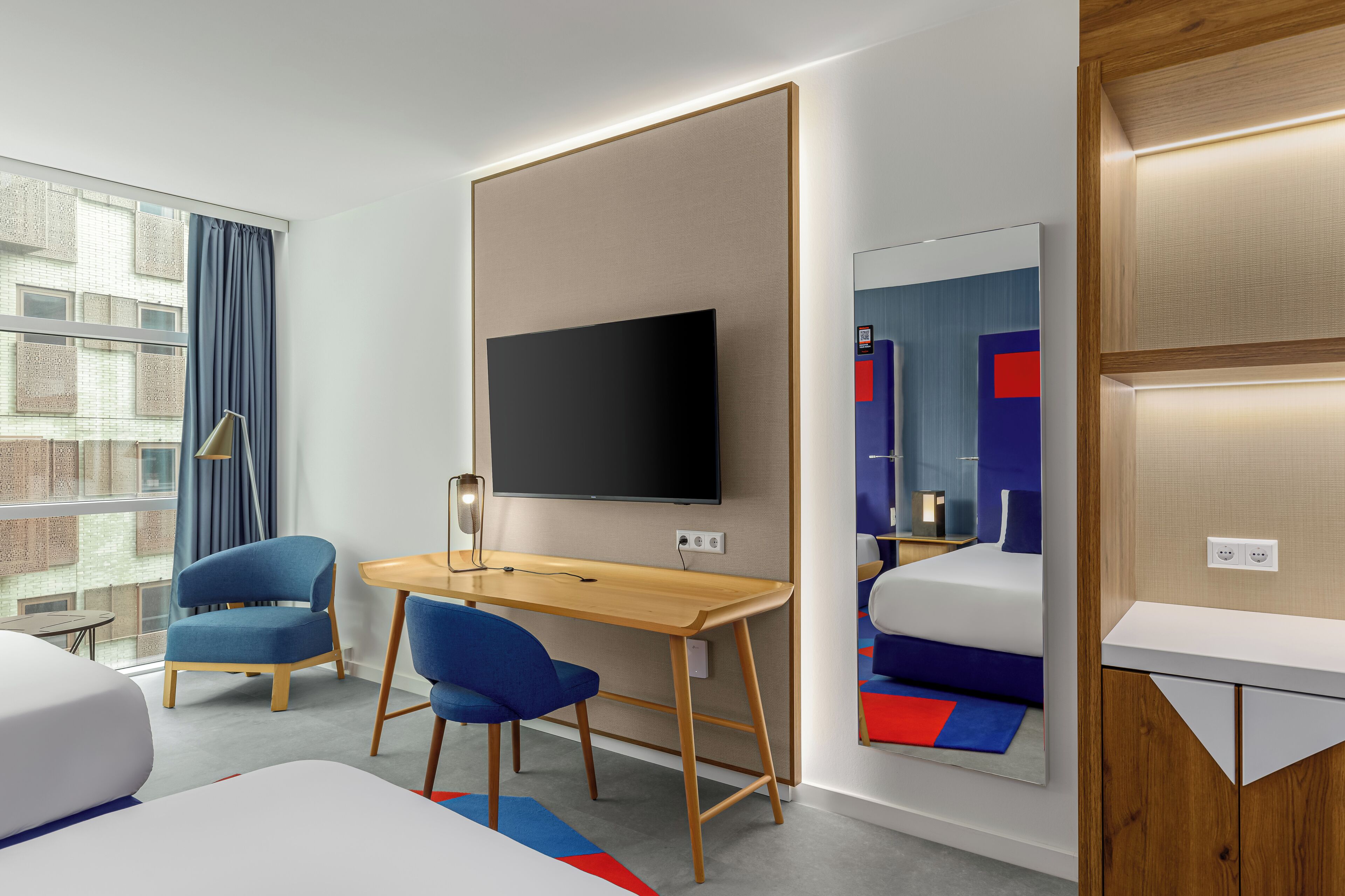 Quarto de hotel moderno com escrivaninha de madeira, televisão de tela plana, espelho grande e cama com tapete colorido.