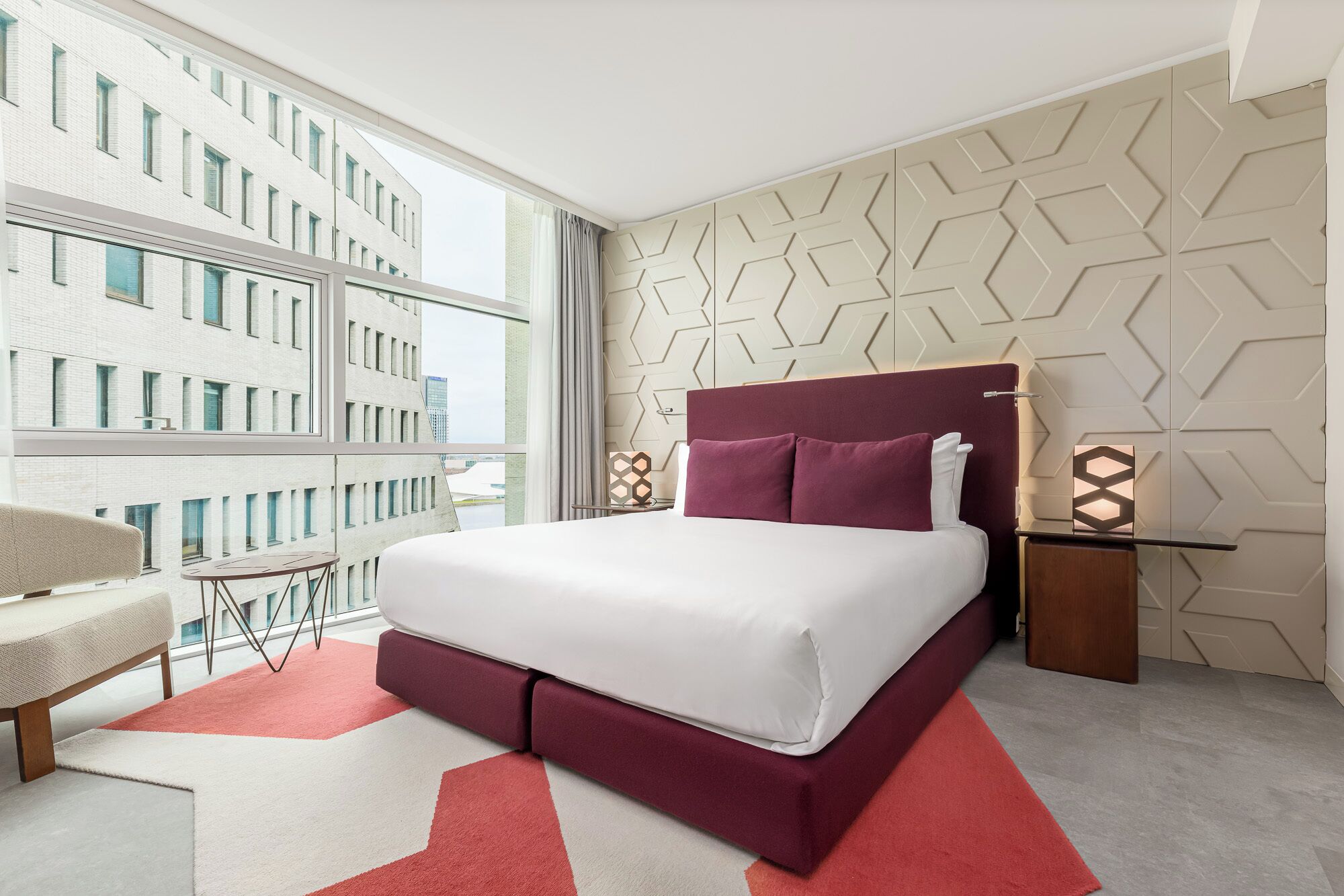 Quarto de hotel moderno com uma cama grande, vista para o horizonte urbano e paredes com padrão geométrico.