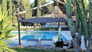 Uma área de piscina com espreguiçadeiras, mesa e estrutura com telhado de palha.
