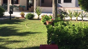 Um edifício de dois andares com telhado de telhas, um alpendre coberto com móveis de exterior e um jardim bem cuidado com diversas plantas e uma caixa de correio vermelha.