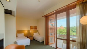 Quarto de hotel com cama, duas mesas de cabeceira, uma cadeira, escrivaninha e varanda com vista para edifícios e vegetação.