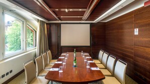 Uma sala de conferências com uma mesa de madeira, cadeiras bege, uma tela de projetor e uma janela com cortinas.
