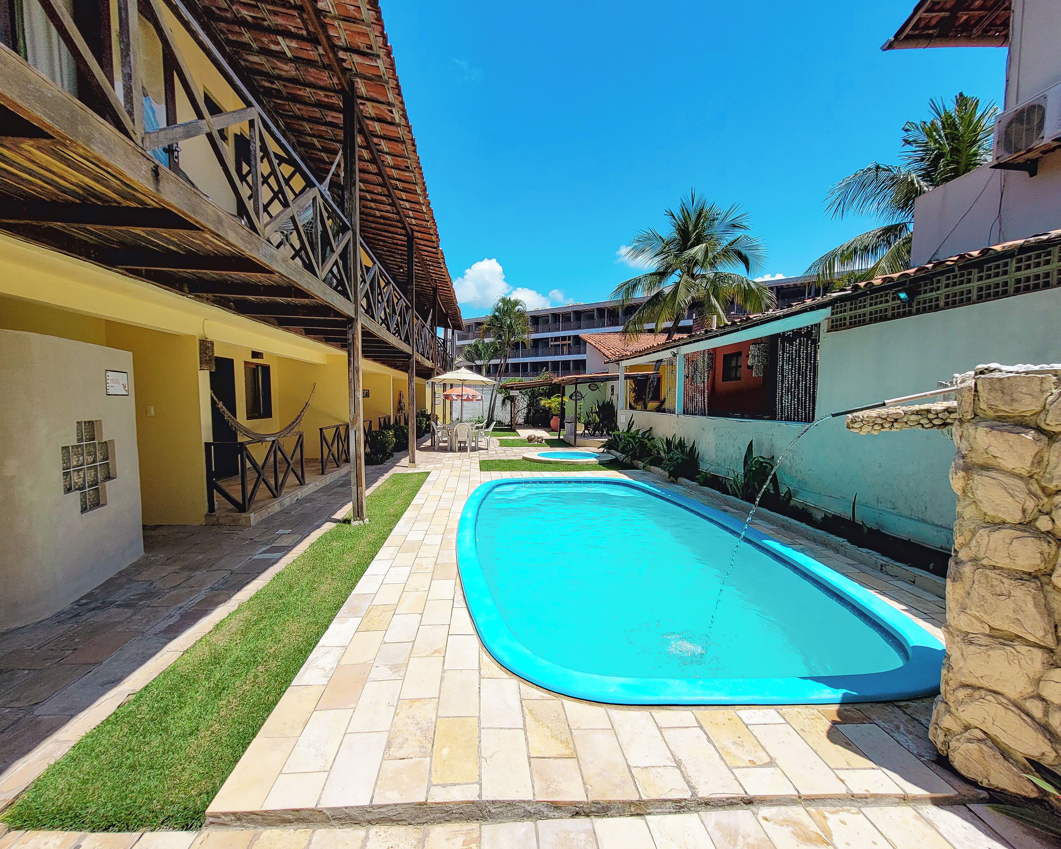 Área de piscina com deck ladrilhado, uma rede e vista para um prédio de vários andares ao fundo.
