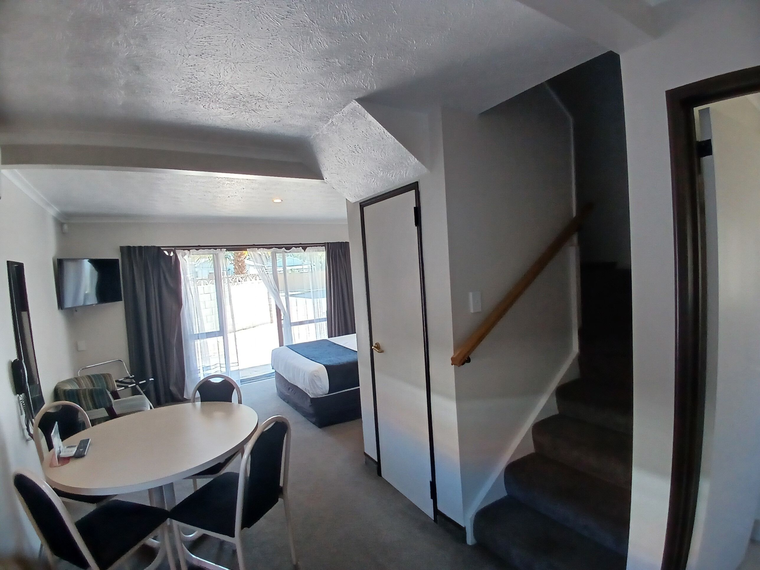 Quarto de hotel compacto com cama, pequena mesa de jantar com cadeiras, televisão na parede e escada para outro andar.