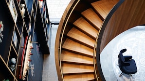 Uma escadaria em espiral com degraus de madeira e guarda-corpo preto, que leva a um patamar com uma cadeira preta e uma parede decorada com diversos objetos.