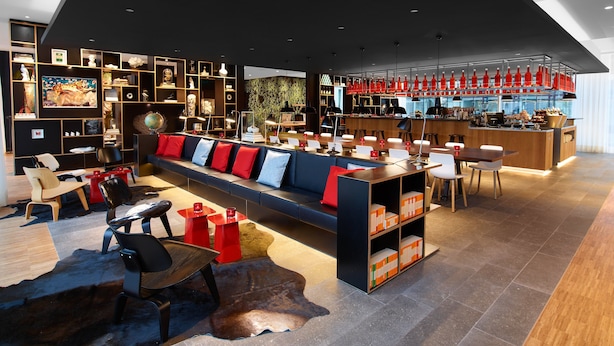 Uma área de estar moderna com um sofá comprido, almofadas vermelhas e azuis, uma mesa de centro de madeira e um bar com luzes pendentes vermelhas.