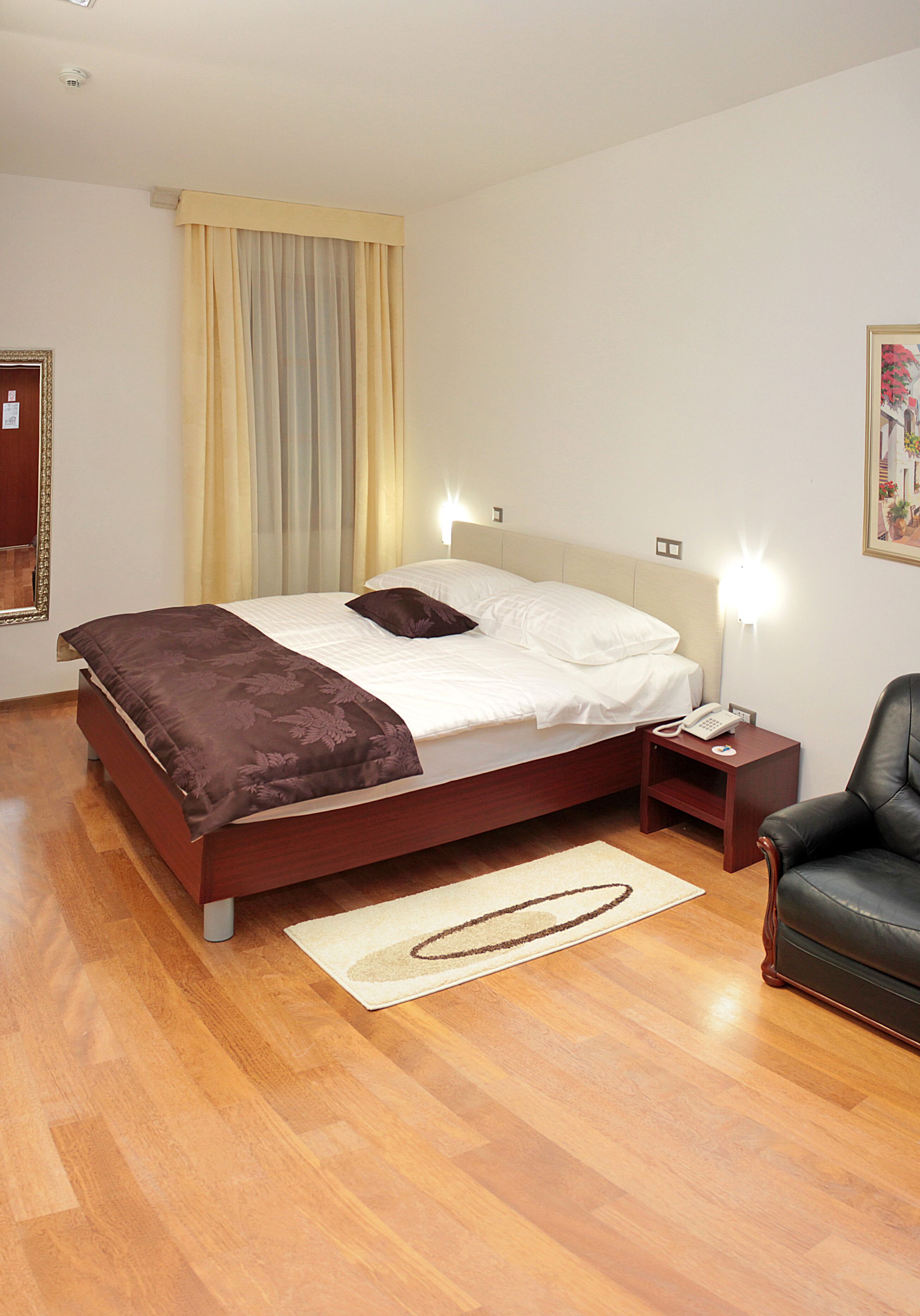 Quarto de hotel com cama de madeira, mesa de cabeceira com telefone, uma cadeira e tapete.