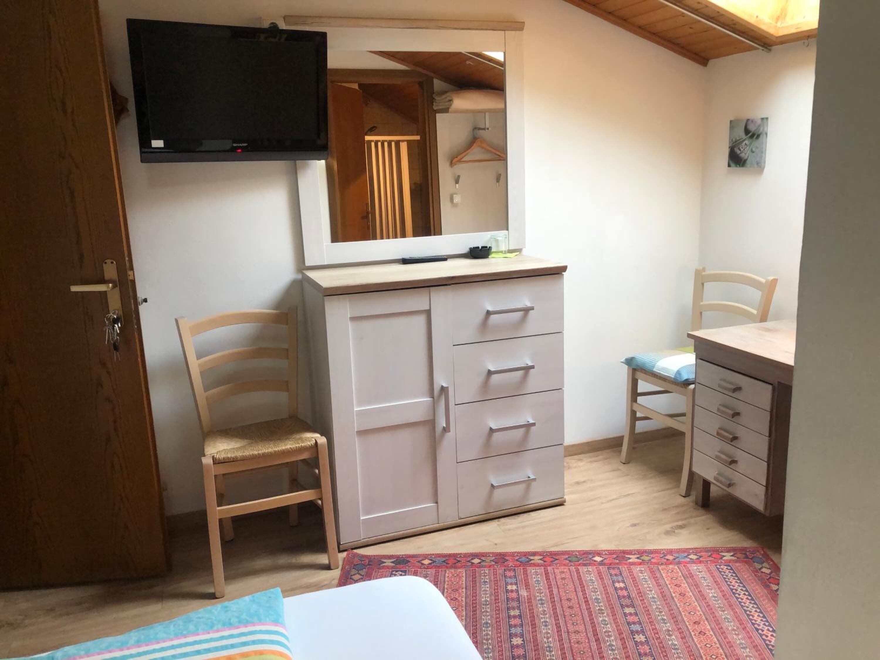 Quarto com cama, escrivaninha, guarda-roupa, cadeira e televisão fixada na parede.