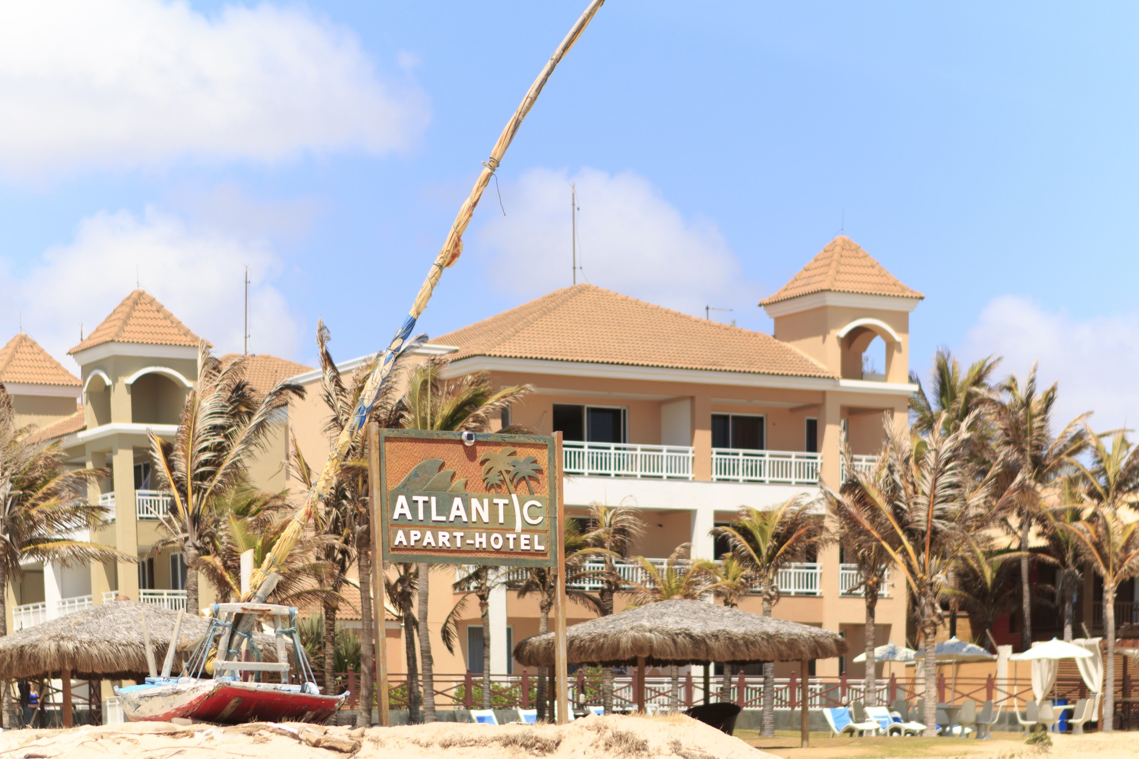 Um hotel à beira-mar com uma placa indicando "ATLANTIC APART-HOTEL", palmeiras, um guarda-sol de palha e um barco.
