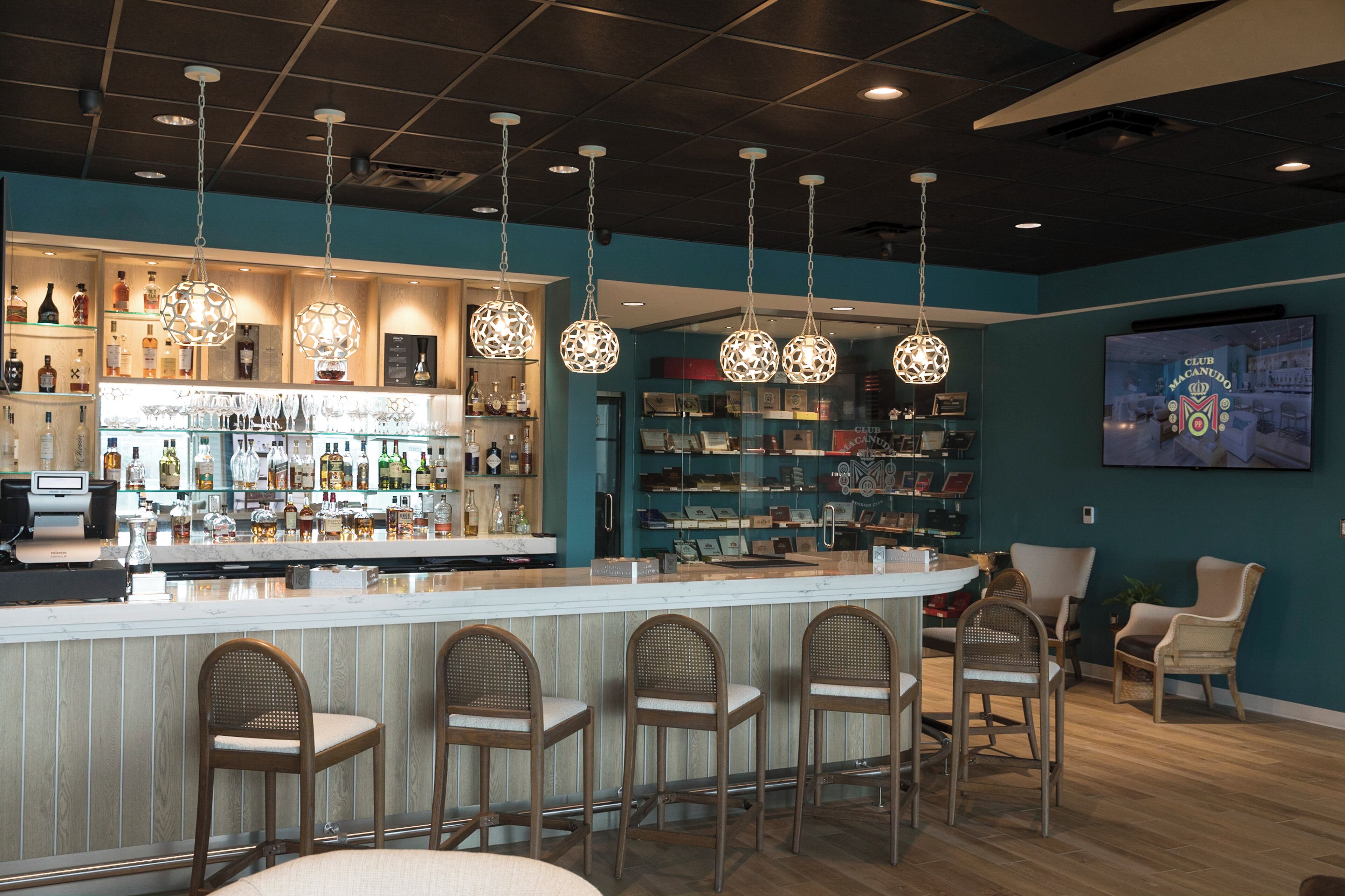 Uma área de bar com balcão, banquetas, luminárias pendentes, prateleiras com garrafas e uma tela de televisão.