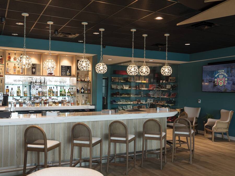 Uma área de bar com balcão, banquetas, luminárias pendentes, prateleiras com garrafas e uma tela de televisão.