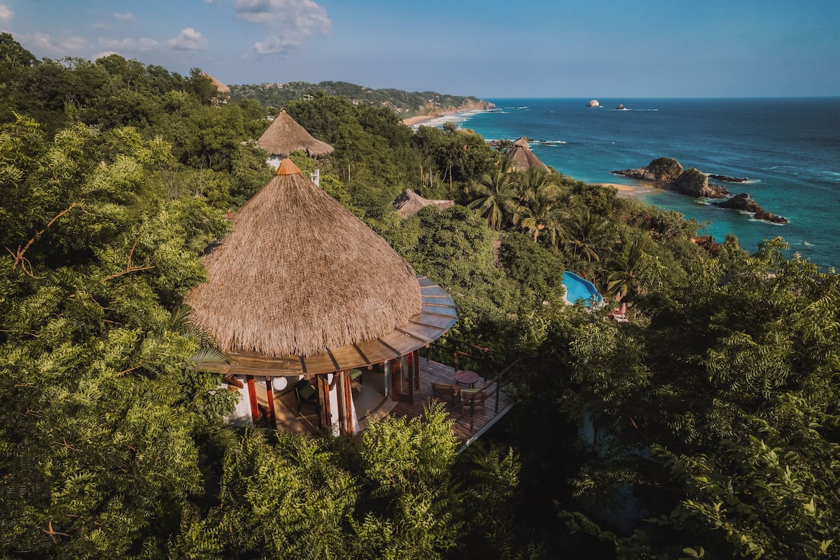 Uma construção com telhado de palha rodeada por folhagem tropical densa, com vista para uma praia e o oceano.