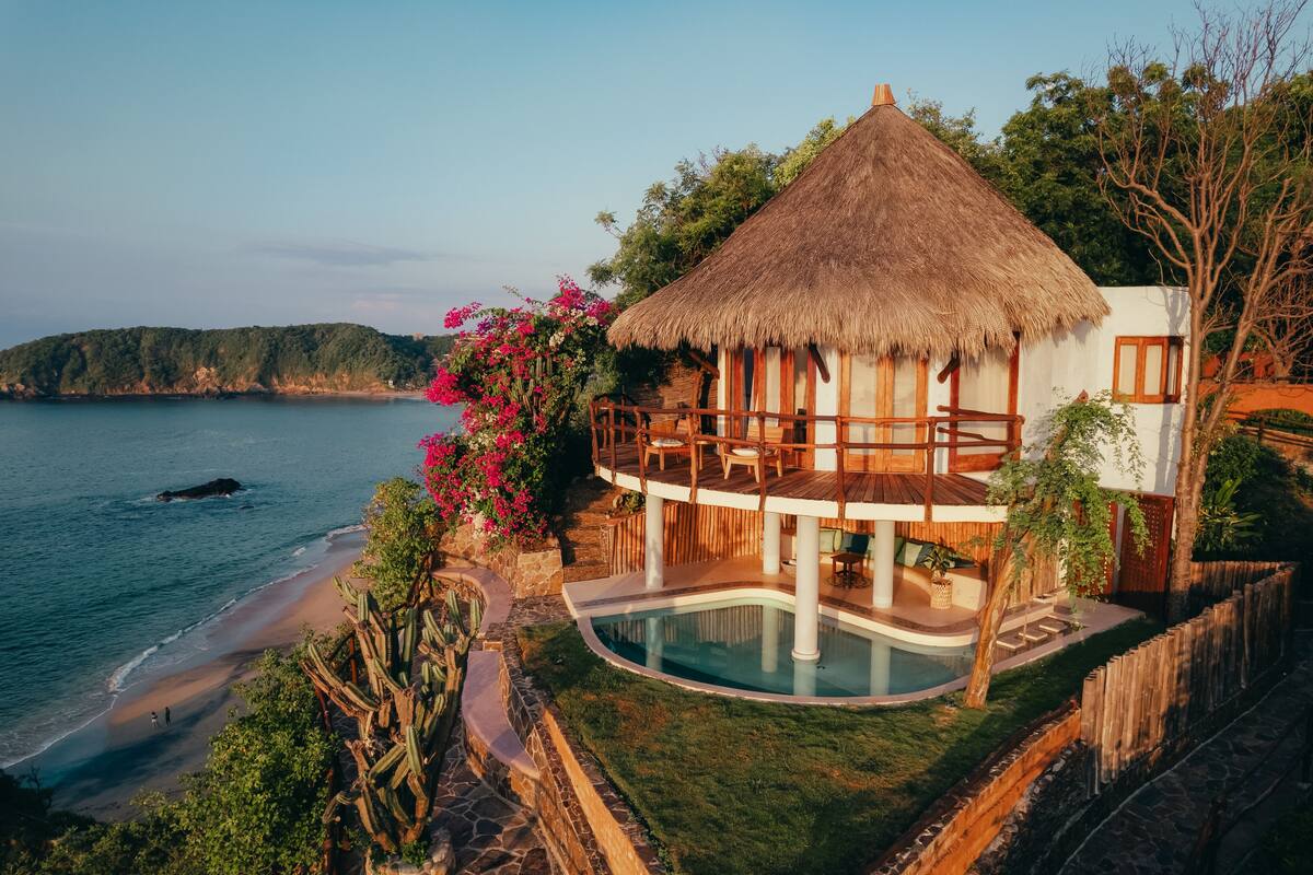 Casa com telhado de palha e piscina, com vista para a praia e o mar.