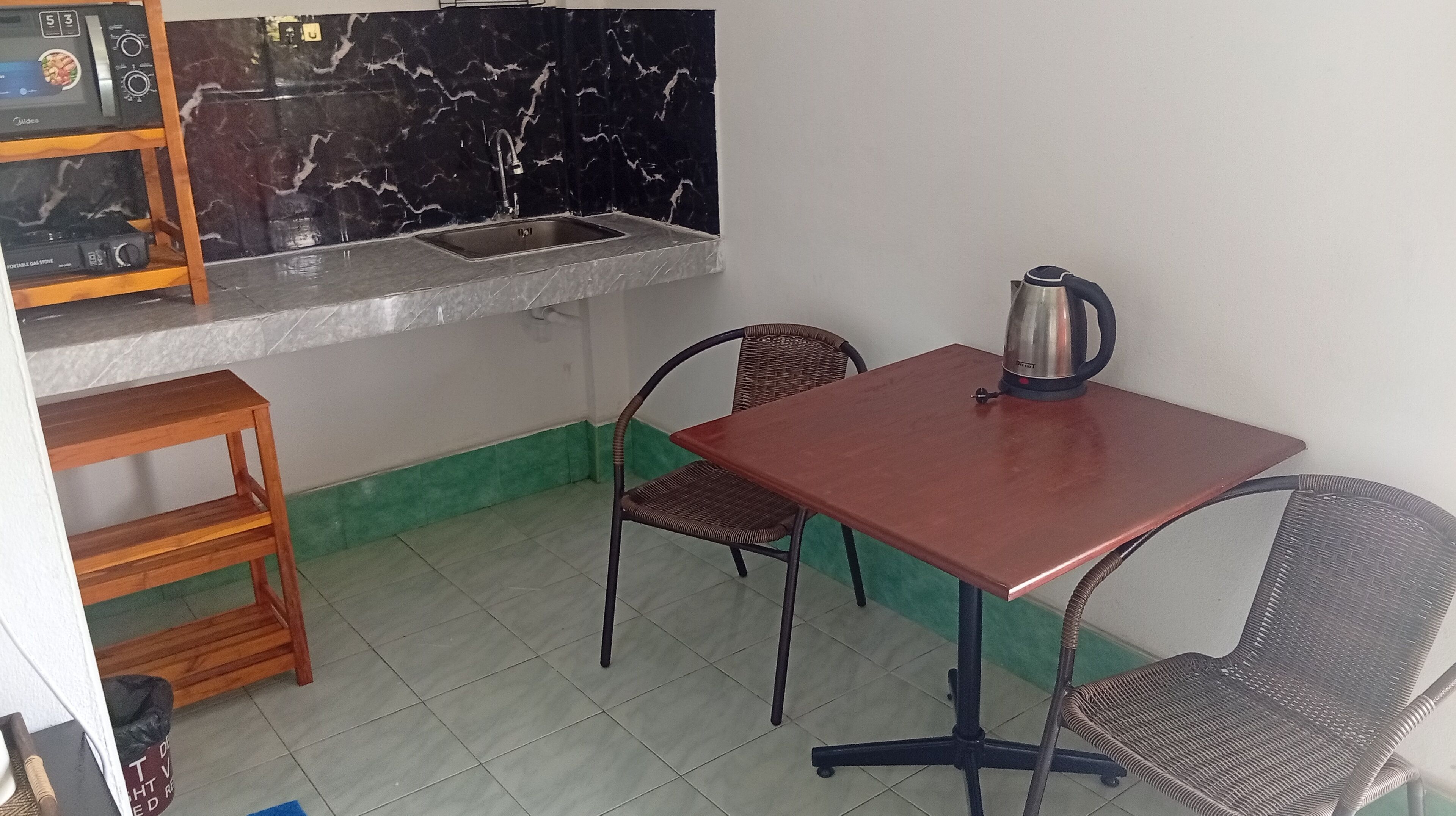 Uma cozinha compacta com pia, fogão a gás portátil e chaleira sobre uma mesa. Há duas cadeiras de vime e um banquinho de madeira.