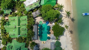 Vista aérea de um resort com vários edifícios com telhados verdes, uma área de piscina central com guarda-sóis e uma praia com um barco.