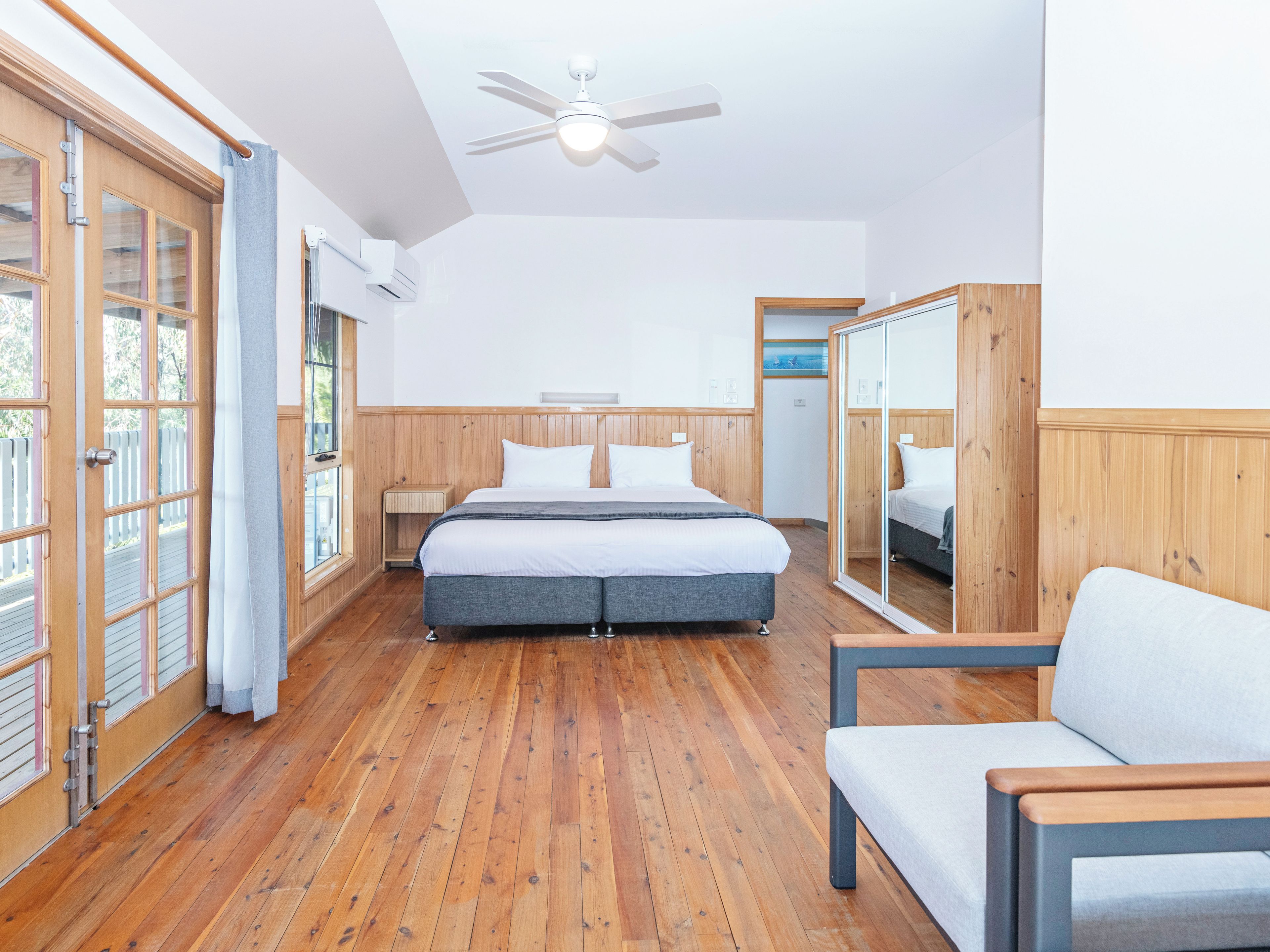 Quarto de hotel com piso de madeira, cama com roupa de cama branca, ventilador de teto, espelho grande e janela com cortinas.