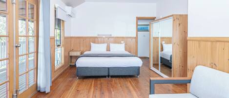 Quarto de hotel com piso de madeira, cama com roupa de cama branca, ventilador de teto, espelho grande e janela com cortinas.