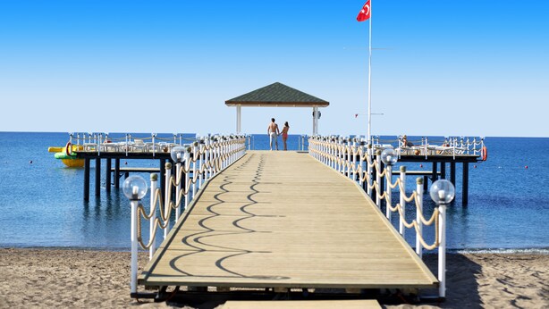 Um píer de madeira que leva a um gazebo numa praia, sob um céu azul límpido.