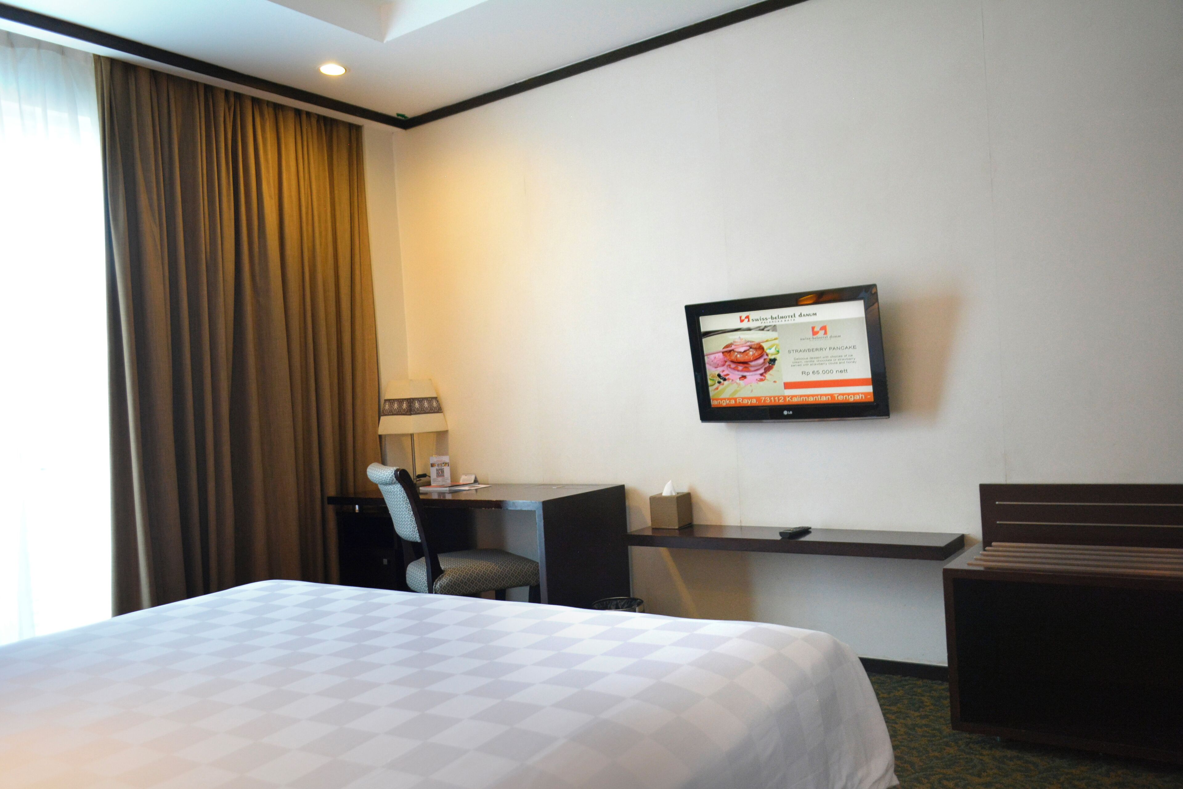 Quarto de hotel com cama, escrivaninha, cadeira, televisão e janela com cortinas.