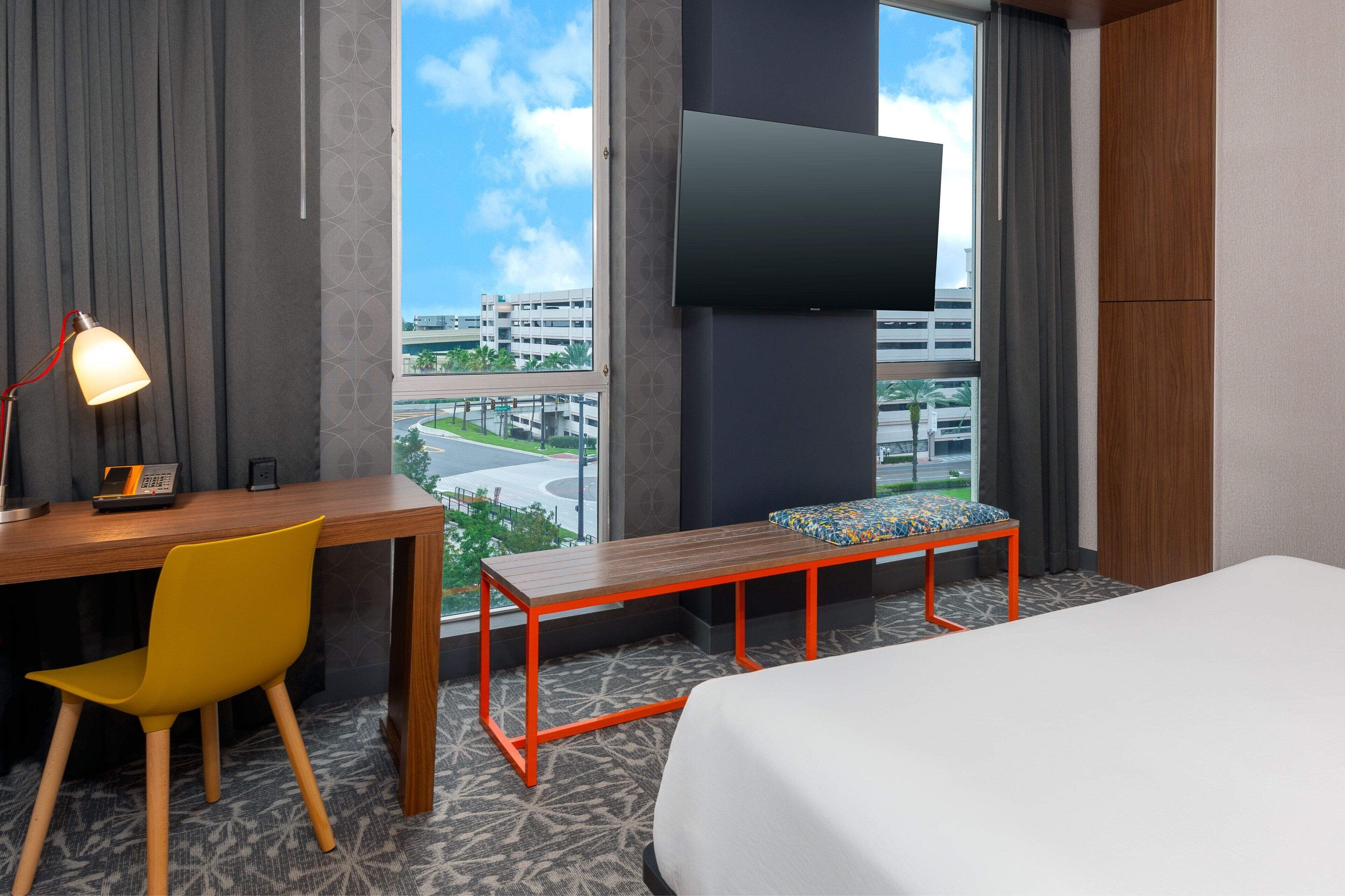 Quarto de hotel com uma cama, uma escrivaninha com luminária, uma cadeira, um banco e uma grande janela com vista para edifícios e uma estrada.