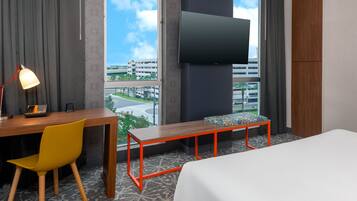 Quarto de hotel com uma cama, uma escrivaninha com luminária, uma cadeira, um banco e uma grande janela com vista para edifícios e uma estrada.