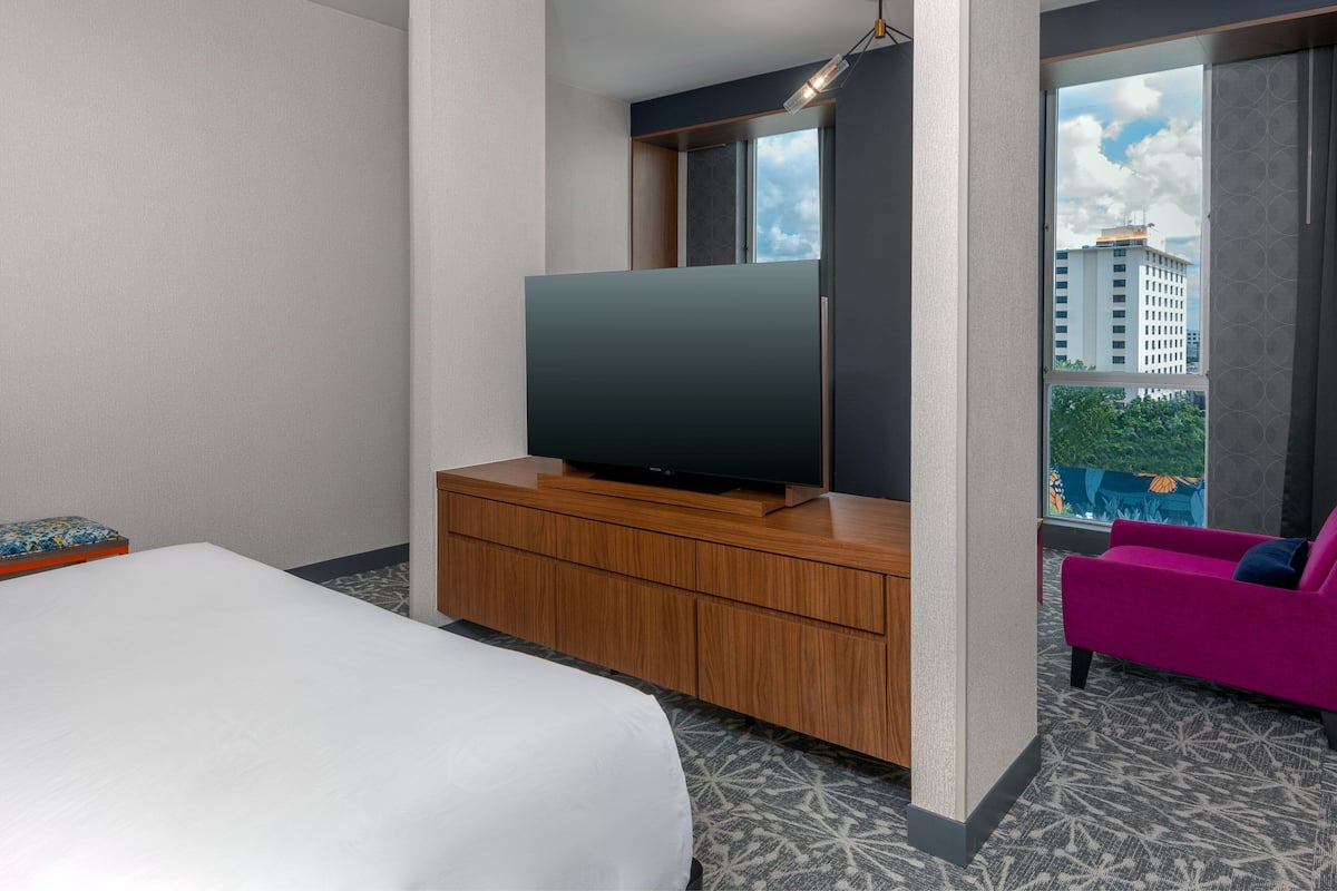 Quarto de hotel com uma cama, uma televisão sobre um móvel de madeira, um sofá rosa e uma janela grande com vista para a cidade.