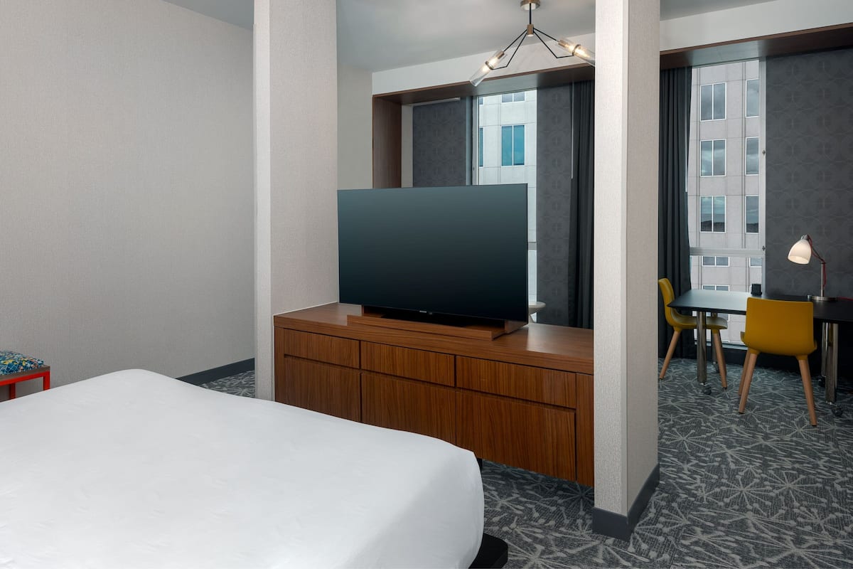 Quarto de hotel com uma cama grande, cómoda de madeira, televisão de ecrã plano, secretária com cadeira e janela com vista para edifícios.