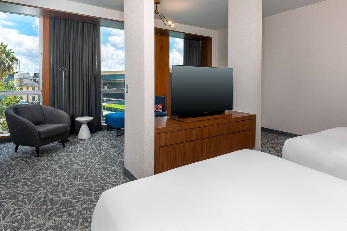 Quarto de hotel com uma grande TV de tela plana sobre um aparador de madeira, uma poltrona cinza, uma pequena mesa lateral branca e duas camas com lençóis brancos.