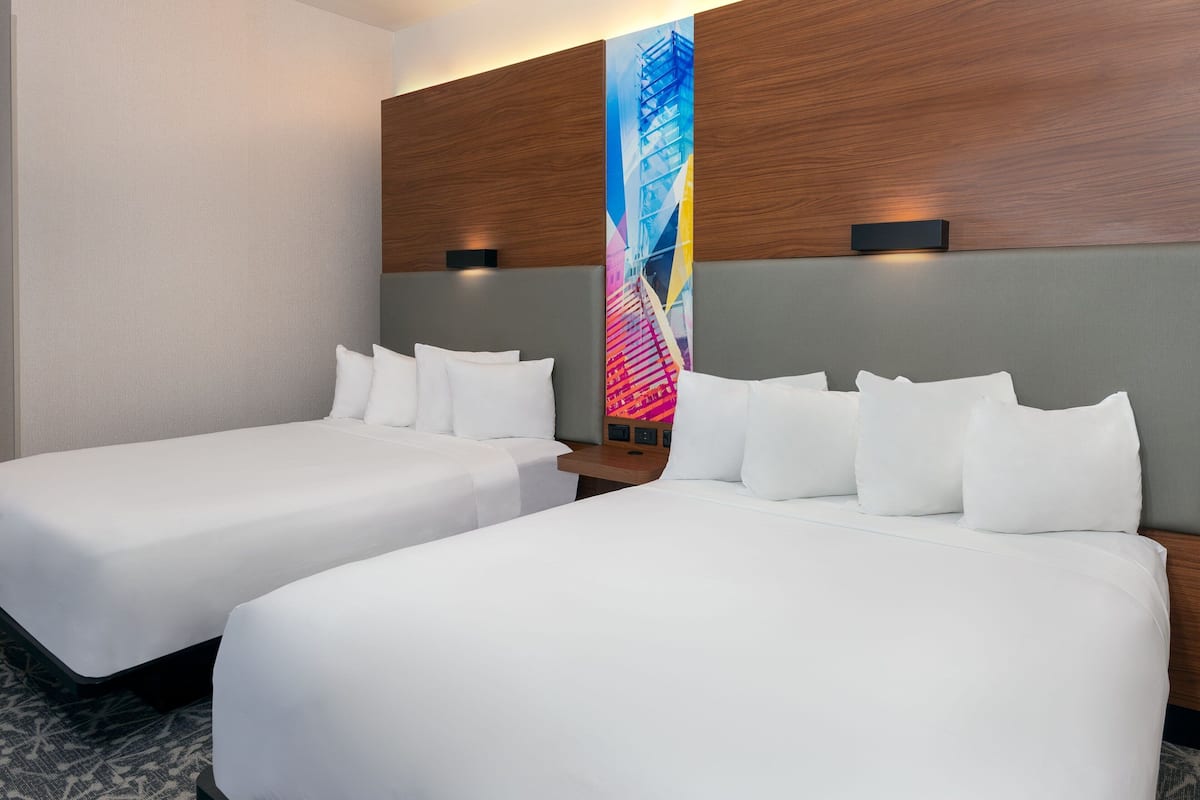 Quarto de hotel com duas camas de solteiro, uma cabeceira de madeira, uma obra de arte grande e colorida e uma mesa pequena.