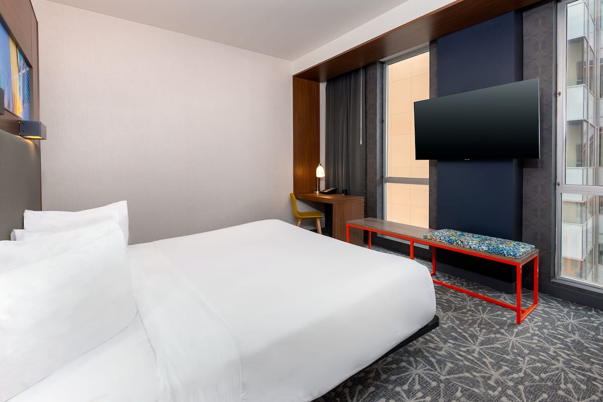 Quarto de hotel com uma cama grande, uma escrivaninha com cadeira, um banco, uma televisão de tela plana e uma janela com vista.