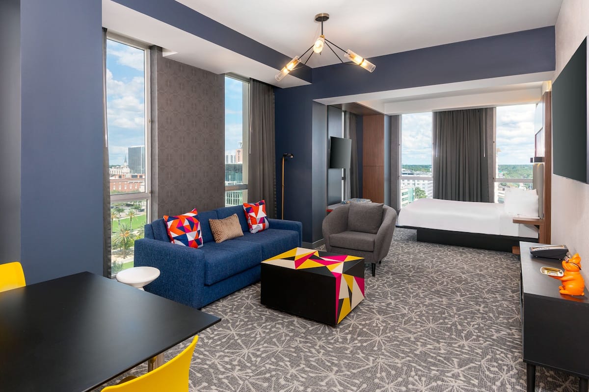 Quarto de hotel moderno com um sofá azul, uma poltrona cinzenta, uma mesa de centro preta e uma cama com cabeceira branca.