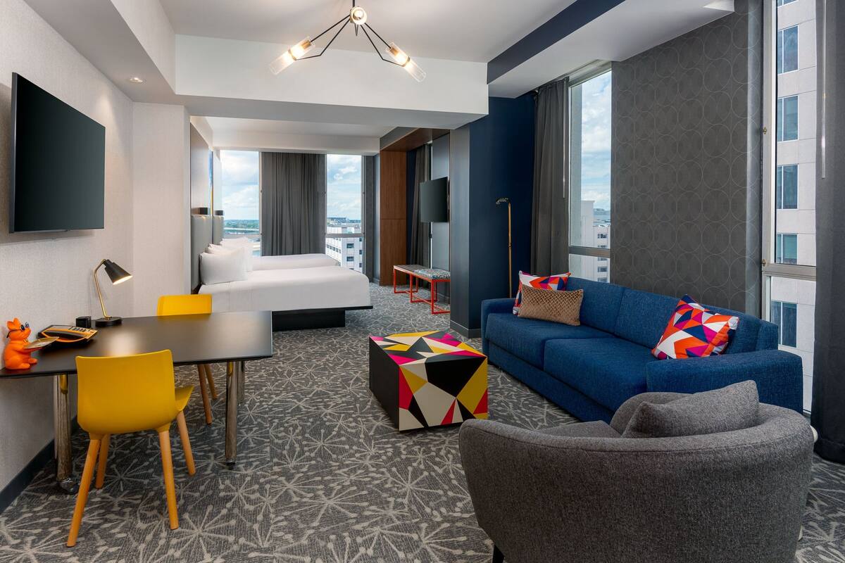Quarto de hotel moderno com uma cama grande, um sofá azul, uma poltrona cinzenta, uma mesinha e uma TV fixada na parede.