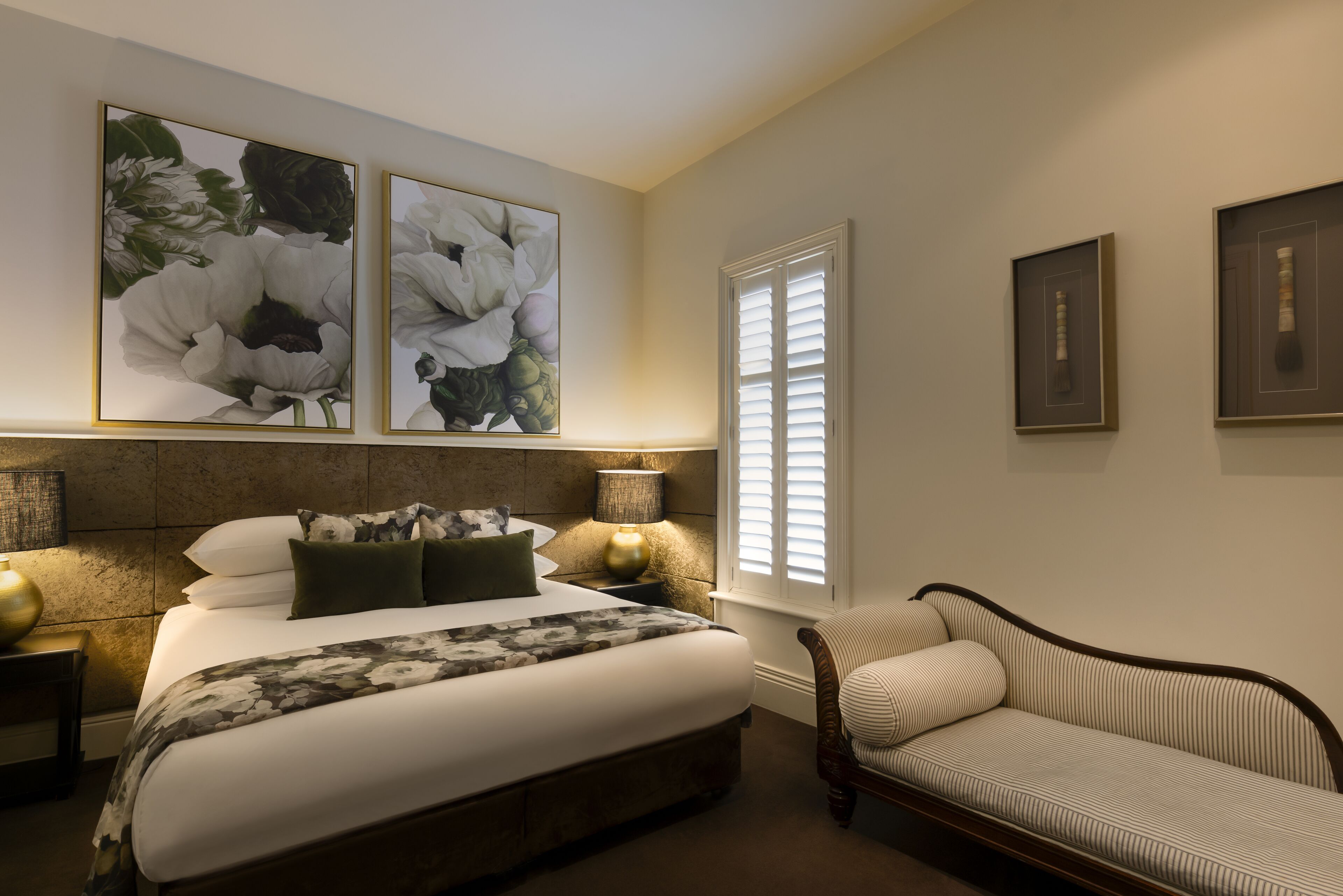 Quarto com cama grande, duas pinturas na parede, abajur, janela com persianas e uma cadeira.