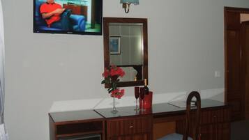 Quarto com televisão mostrando um homem de camisa vermelha, uma escrivaninha de madeira com um espelho, um vaso com flores vermelhas e uma cadeira.