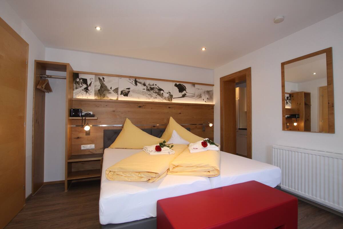 Quarto de hotel bem arrumado, com cabeceira de madeira, cama grande com roupa de cama e almofadas amarelas, um banco vermelho e um espelho fixado na parede.