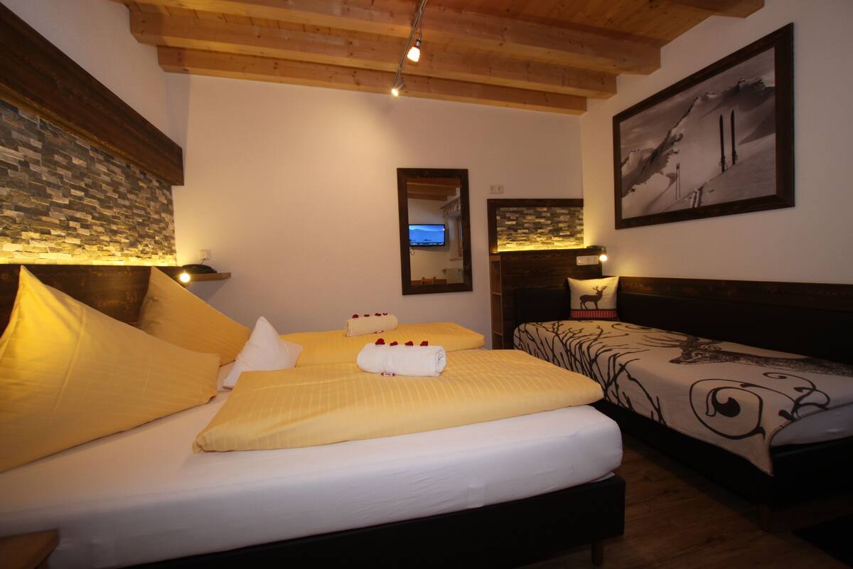 Quarto de hotel com uma cama grande, uma cama menor, teto de madeira, parede de pedra, televisão montada e uma moldura com um quadro na parede.