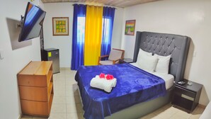 Quarto de hotel com cama, televisão, escrivaninha, cadeira e mesa de cabeceira.