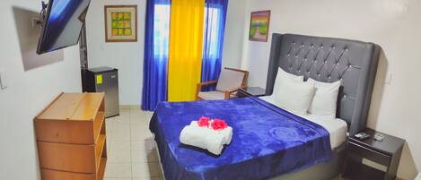 Quarto de hotel com cama, televisão, escrivaninha, cadeira e mesa de cabeceira.