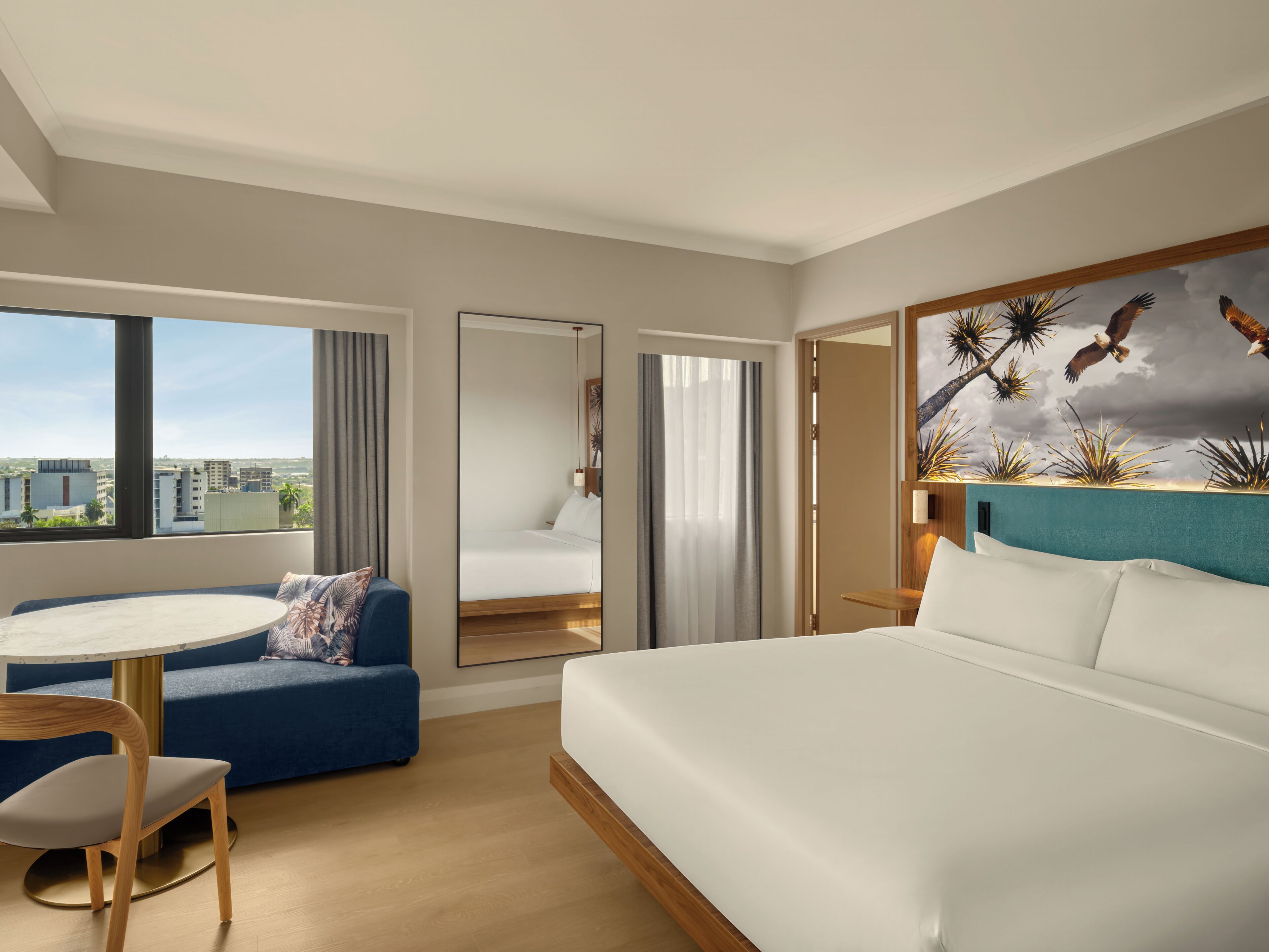 Quarto de hotel com cama, área de estar com mesa e uma grande janela com vista para a cidade.