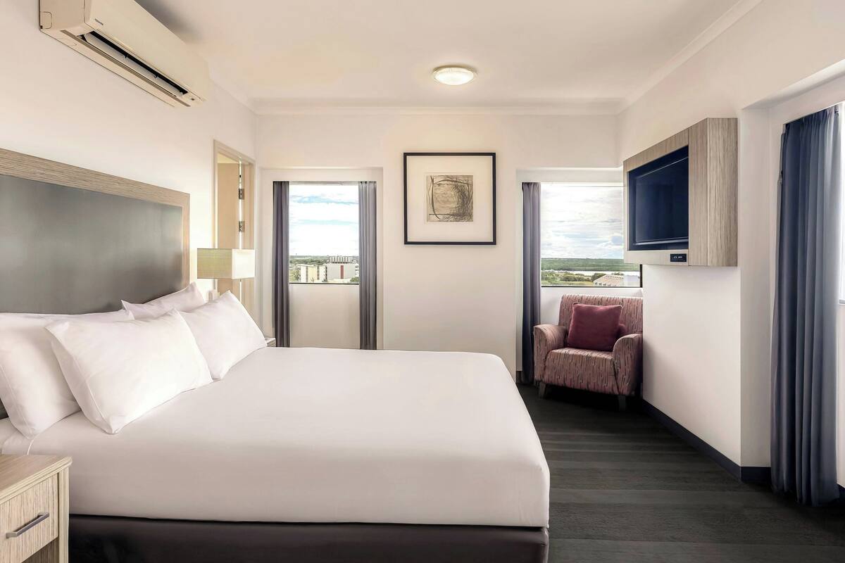Quarto de hotel com uma cama grande, televisão fixada na parede, uma cadeira e janela com vista para a cidade.