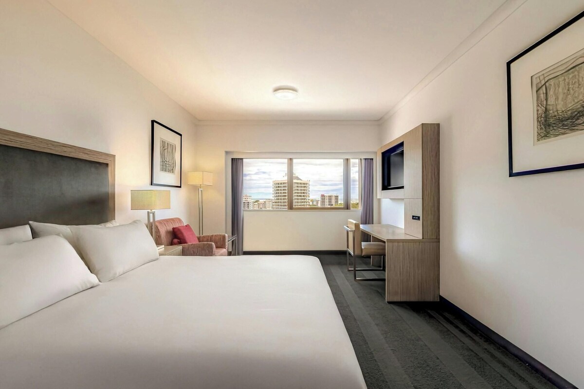 Quarto de hotel com uma cama grande, uma mesa de trabalho, uma cadeira, uma TV e uma janela com vista para a cidade.