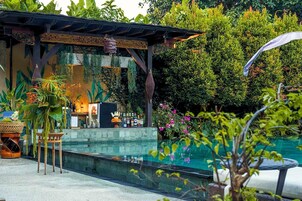 Uma área de estar coberta ao ar livre, com piscina, rodeada por vegetação exuberante e diversas plantas.