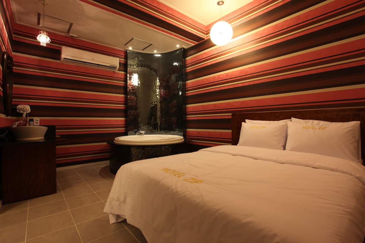 Quarto de hotel com paredes listradas, uma banheira redonda, uma cama com almofadas rotuladas "HOTEL ZIP" e uma pia com espelho.