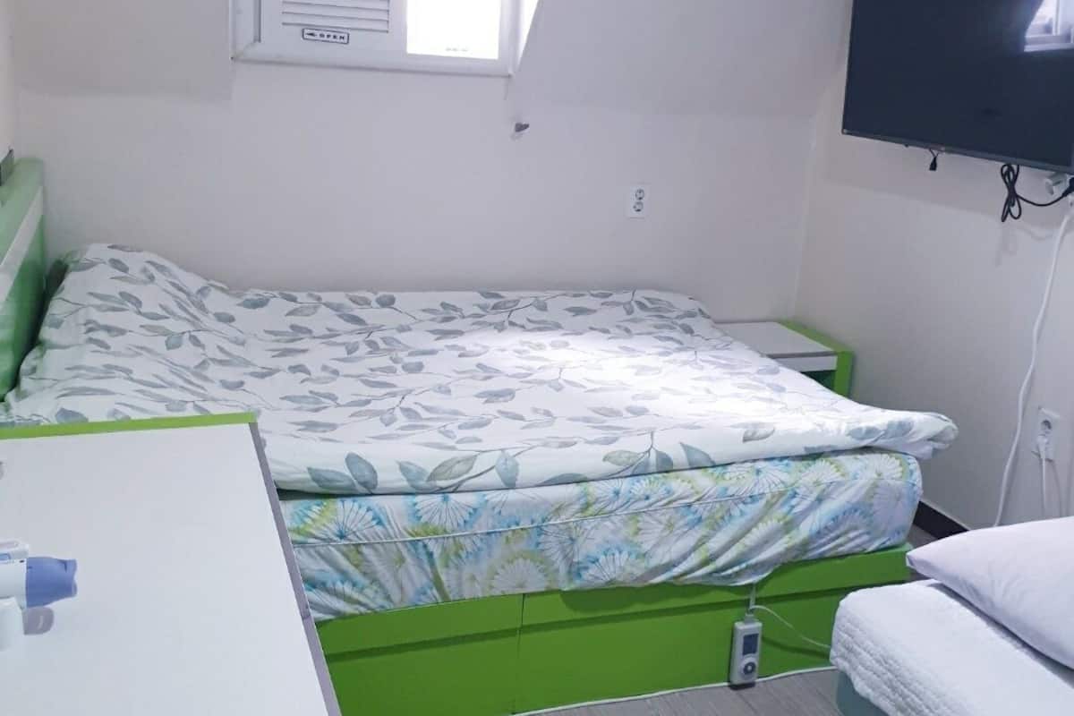 Um apartamento pequeno e de um único cômodo, com teto inclinado, cama com colcha florida, escrivaninha com computador e TV na parede.