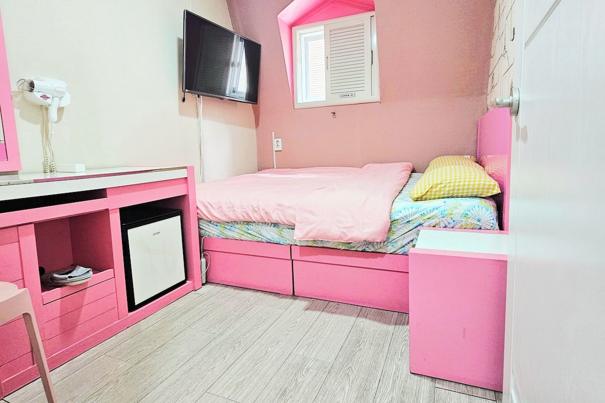 Quarto com cama rosa, televisão na parede e escrivaninha com computador.