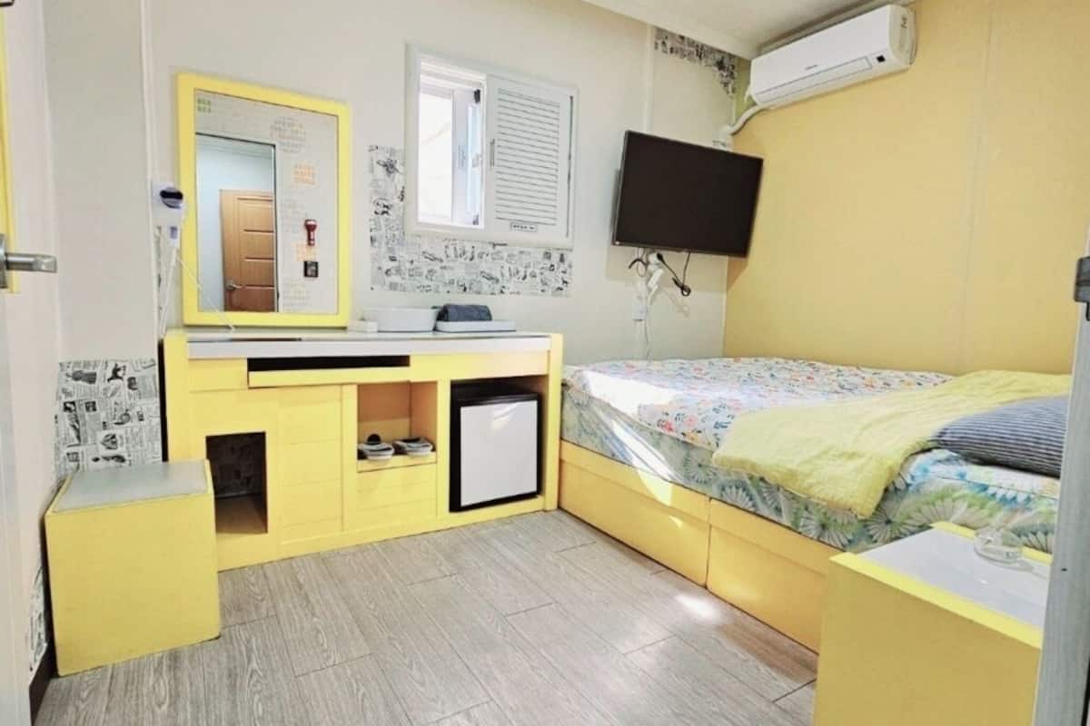 Quarto compacto com uma cama, uma escrivaninha pequena, um espelho, uma TV e um ar condicionado.