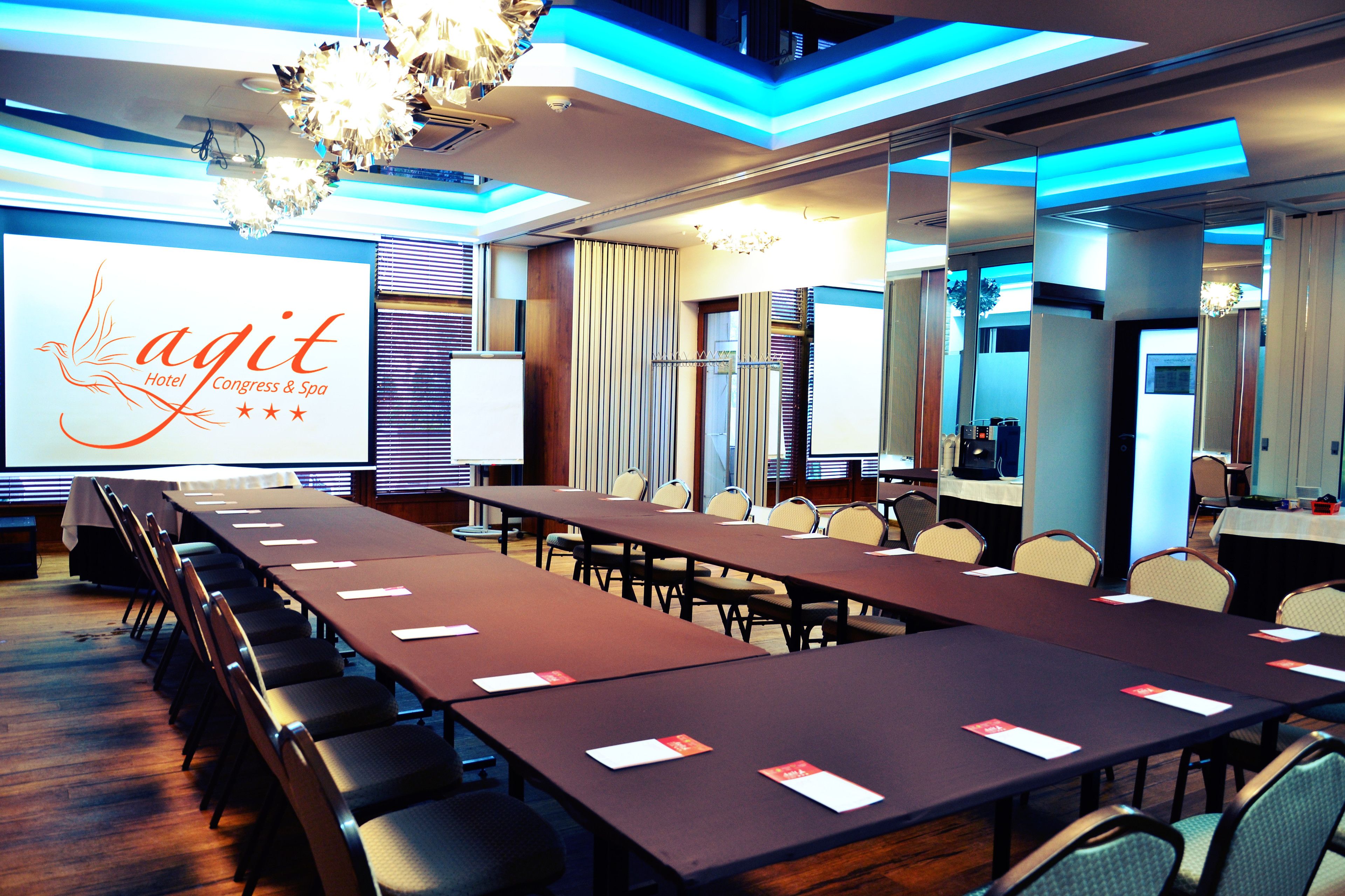 Sala de conferências com mesas compridas, cadeiras e uma grande tela exibindo o nome do hotel.