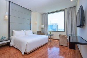 Quarto de hotel com uma cama grande, escrivaninha, cadeira, televisão e janela com cortinas.