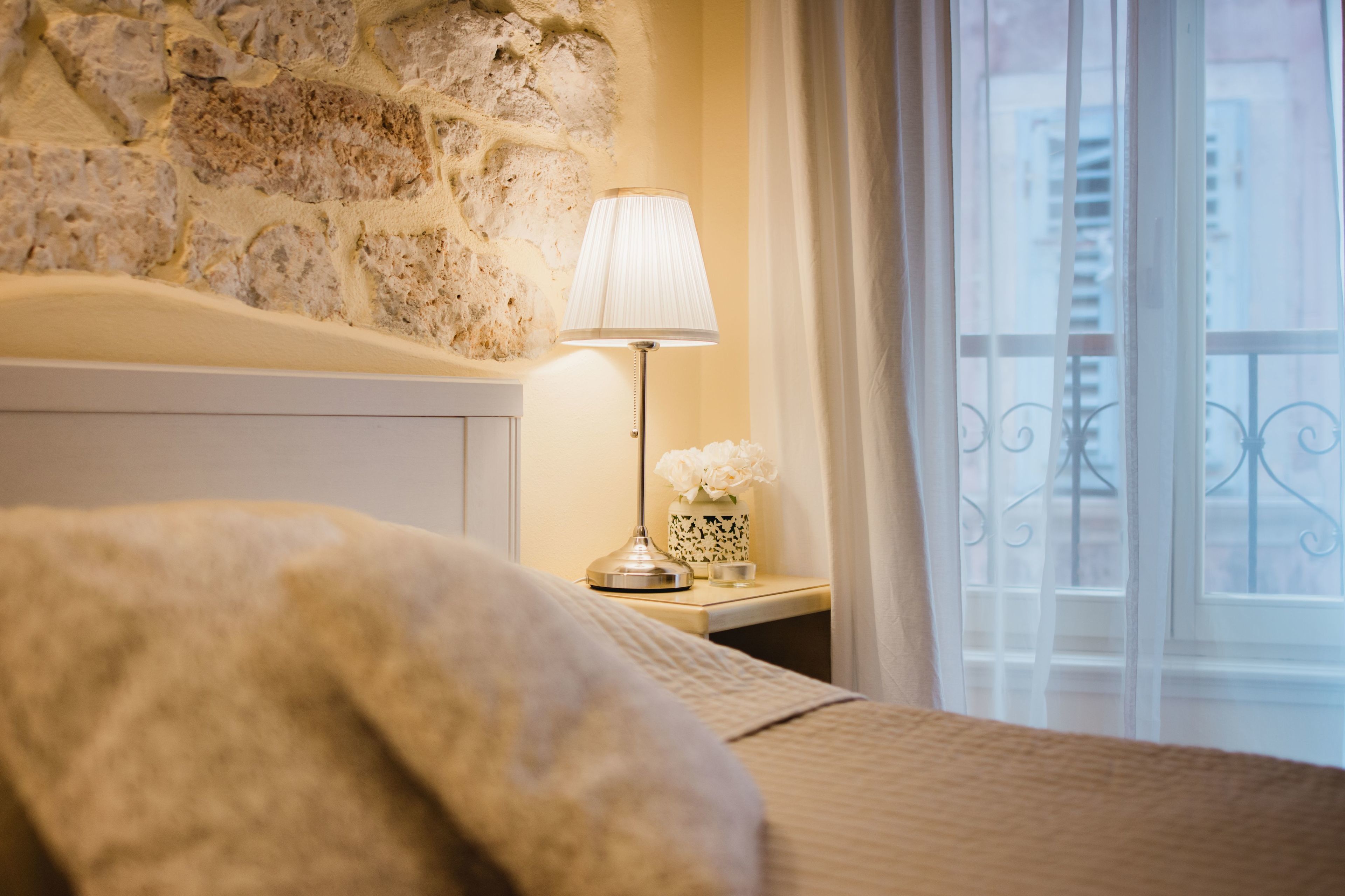 Quarto com parede de pedra, abajur, vaso com flores e vista para uma varanda com grades de ferro.